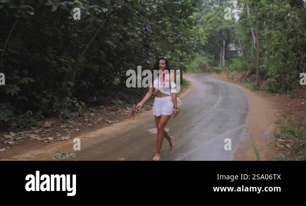 A stylish woman walks barefoot along a wet rural road in Sri Lanka, wearing a white outfit and a ...