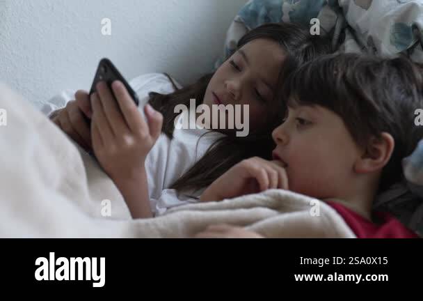 Small siblings staring at cellphone device in bed, sister sharing phone ...