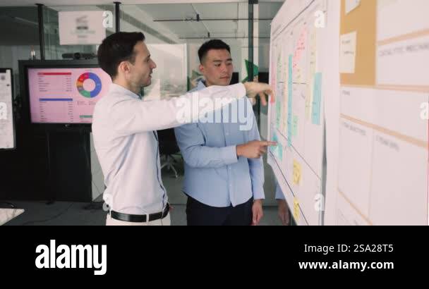 Two diverse male colleagues discussing tasking strategy at Kanban board ...