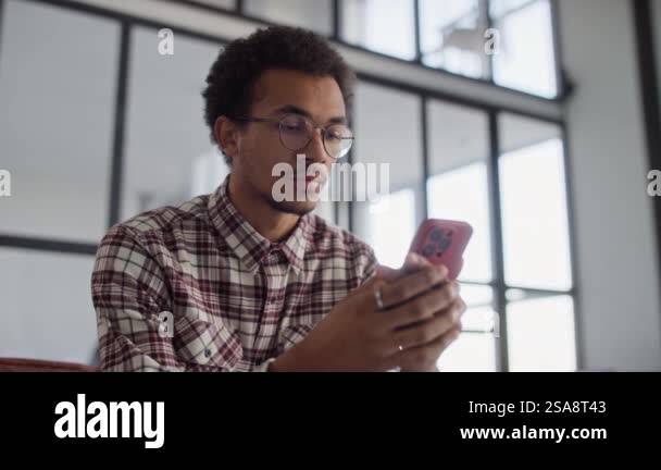 A young man in a plaid shirt and glasses smiles while texting on his ...