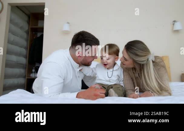 Mother and father lie on the bed and their little baby sits between ...