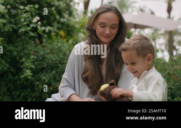 Son plays with small toy as mother observes with warm smile, creating ...