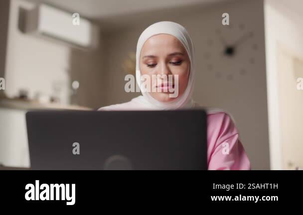 At kitchen counter, Muslim woman in white hijab types email on laptop ...
