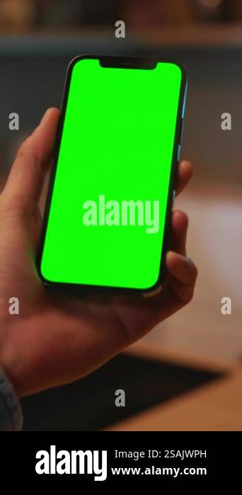 Mans left hand holiding smartphone with green screen in vertical position isolated on blurred ...