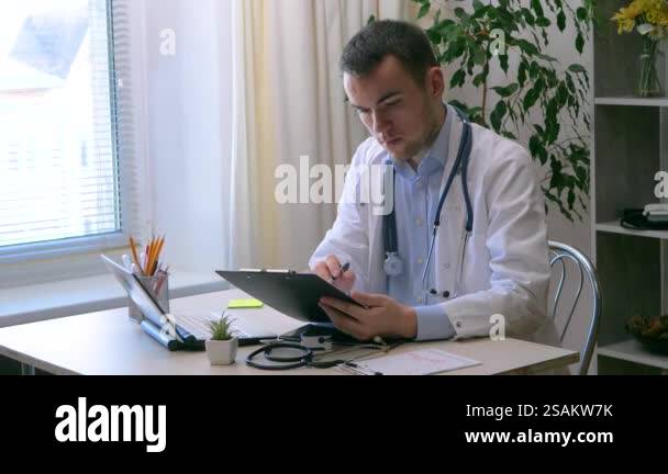 A young intern doctor works in an office, reads a patient's medical ...