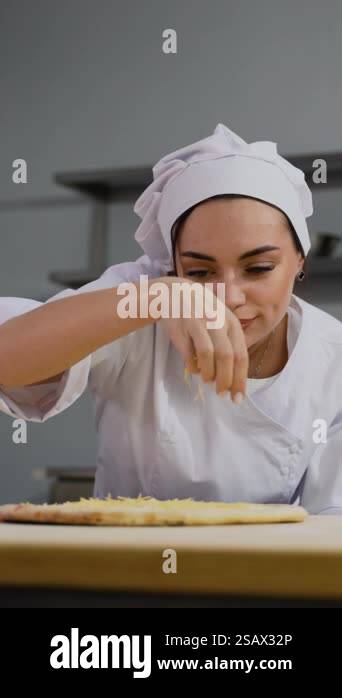 A chef in a white uniform adding shredded cheese to a pizza dough base ...