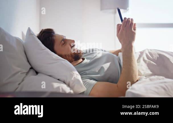 Happy guy looking smartphone screen in bed closeup. Sleepy male read ...
