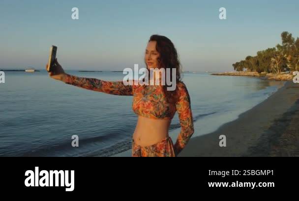 A cheerful woman in a vibrant swimsuit takes a joyful sunset selfie on the beach, capturing ...