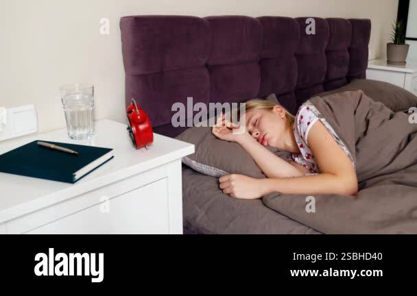 A young girl wakes up from the ringing of a red alarm clock, lazily ...