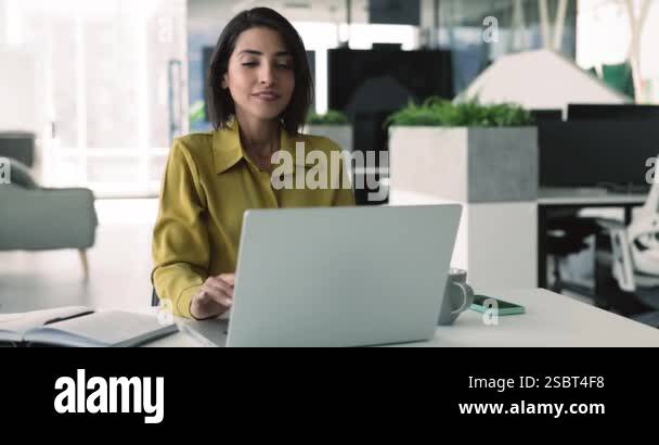 Smiling Brazilian woman seated at desk in office, leaning back with her hands behind head, take ...