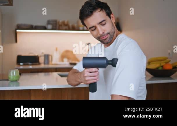 Man massaging shoulder electronic gun in modern kitchen after workout ...