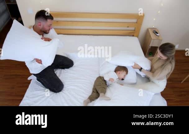 Loving parents sitting on the bed hit their little baby with pillows ...
