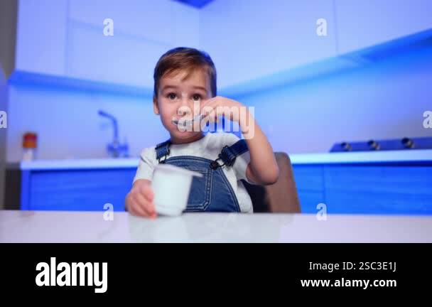 Lovely toddler eating yogurt with a spoon. Kid takes a cup to his mouth ...