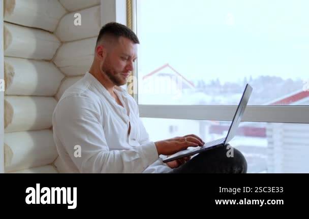 Bearded man sitting at the window holds a laptop on the knee. Freelance ...