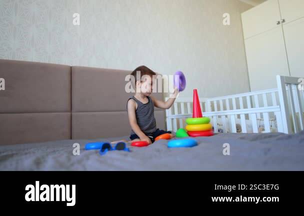 Male kid sits on the bed playing with a pyramid. Baby boy plays with a ...