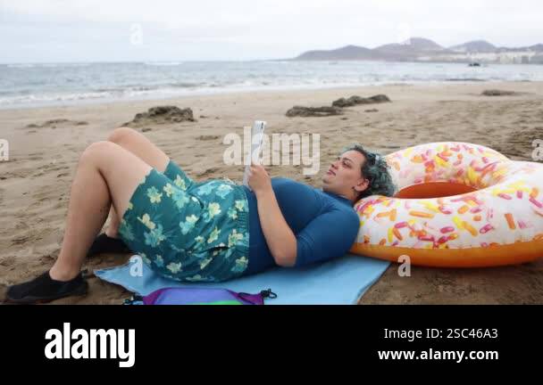 A trans man relaxes on a sandy beach, leaning on a colorful float ...