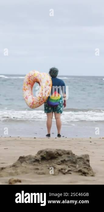 A joyful trans man stands on a sandy beach, holding a colorful ...