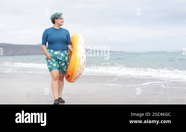 A trans man enjoys a beach vacation, carrying a colorful ring float ...