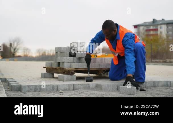 A construction worker in a safety vest and gloves carefully lays paving ...