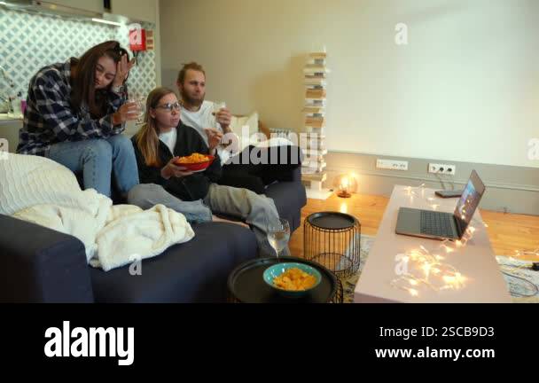 Friends relax on a couch, enjoying snacks and a movie on a laptop in a ...