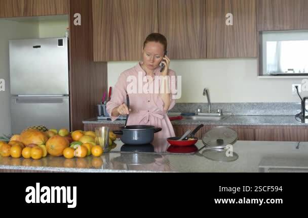 A woman in the kitchen multitasks by talking on a cell phone while ...