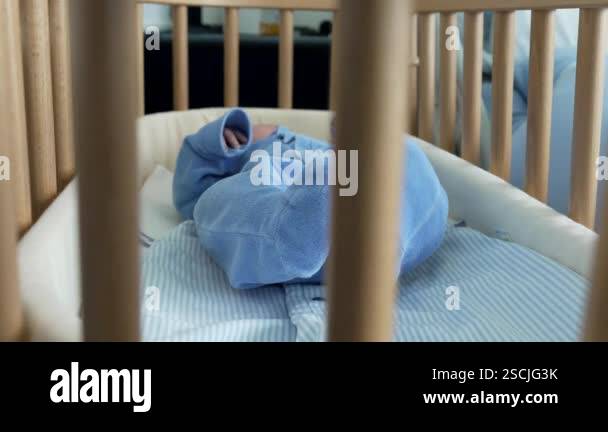 Newborn baby in a blue outfit resting in a wooden crib, viewed through ...