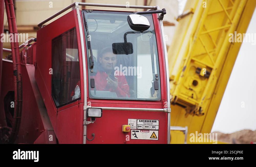 Mature man in red uniform and cap sits in red crane cabin with walkie ...