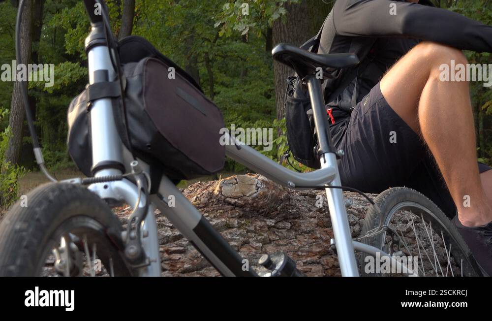 A young handsome cyclist sits on a log next to his bike in a forest and ...