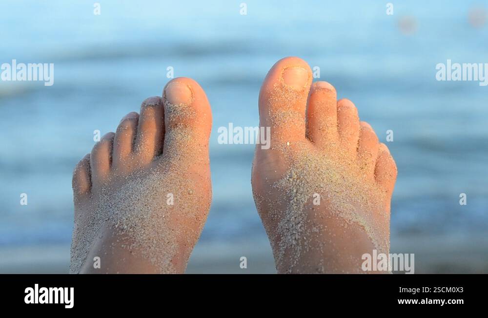 Two feet strewn with sand in front of waves of sea in light of sunset ...