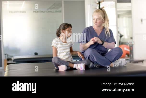 Physical therapist in scrubs helping young girl with cerebral palsy with disability in rehab ...