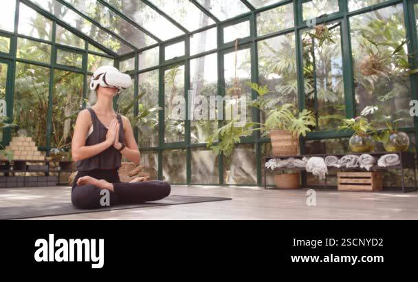 Practicing yoga, woman wearing VR headset in glass house, meditating on ...