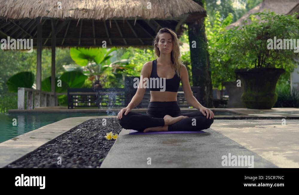 low shot calm woman sitting in lotus pose outside with tropical hut on ...