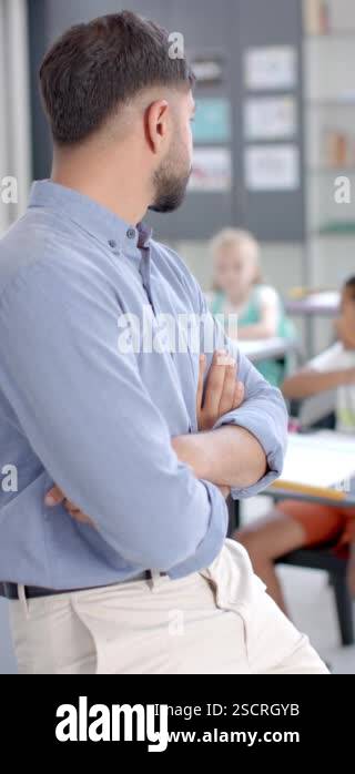 Vertical video: In school, male teacher standing with arms crossed in classroom. Education ...