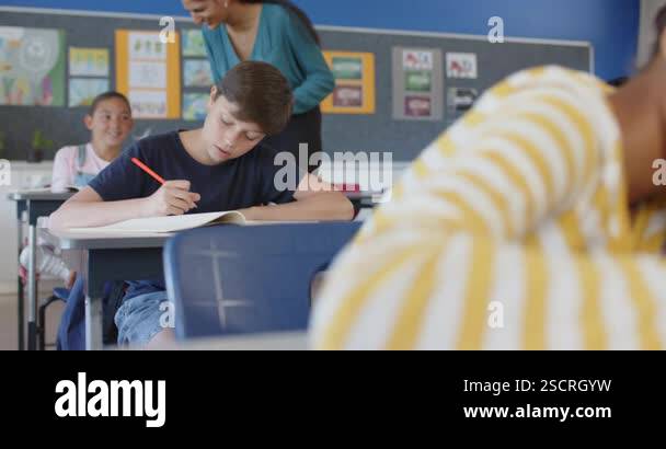 Smiling boy writing in notebook in school classroom with teacher in ...