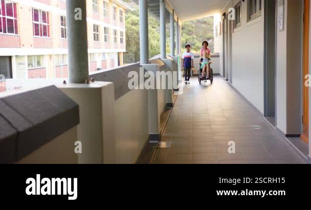 Pushing wheelchair, two girl students helping friend in school hallway ...