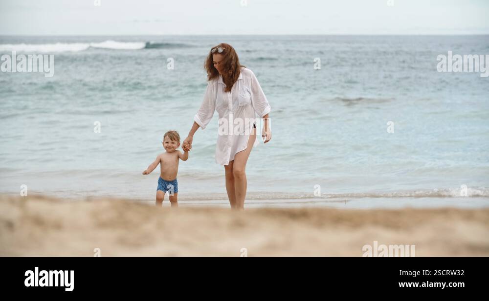 Cute expressive baby making careful steps on shore of ocean, happily ...