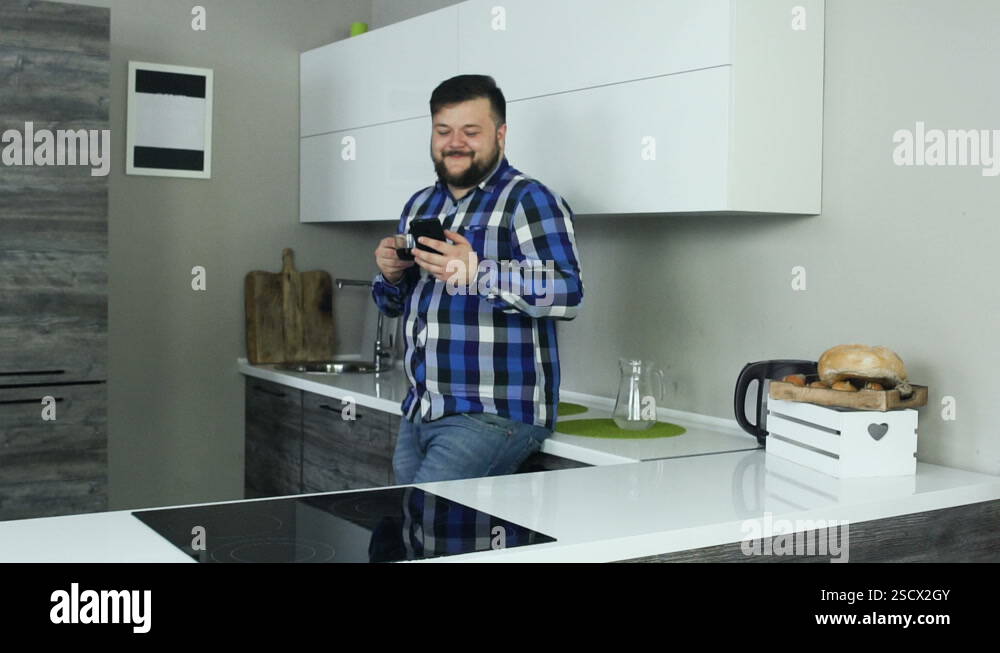 A stout man stands in the kitchen and laughs, looking at the phone ...