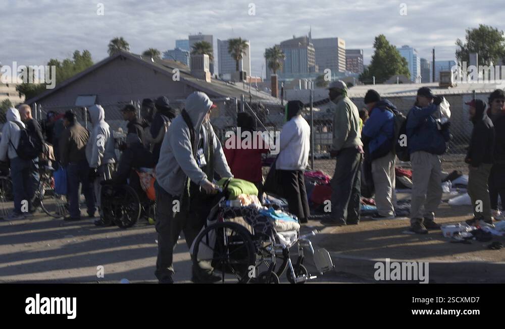 A homeless man pushes a wheelchair past a line of other homeless people ...