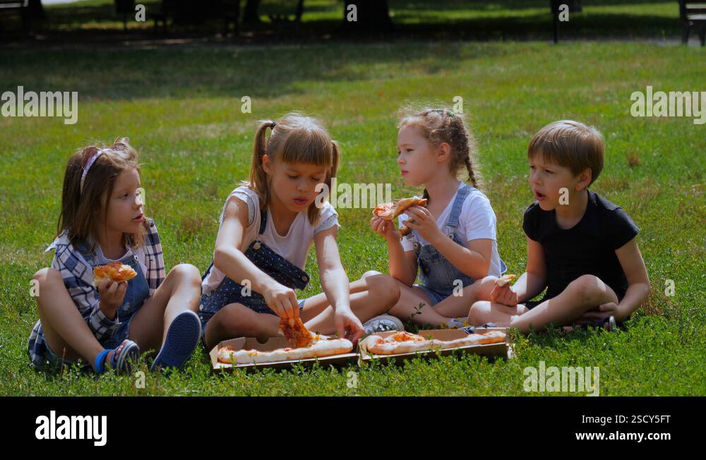 Group of children friends picnic eat pizza sit on park grass chewing ...