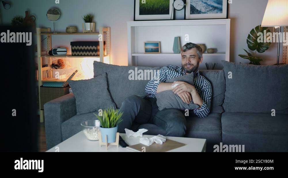 Sensitive man watching sad movie on TV at home wiping eyes with paper ...