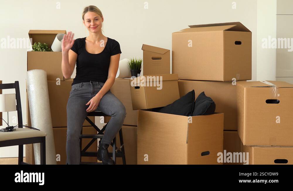 A friendly moving woman sits on a chair in an empty apartment and waves ...