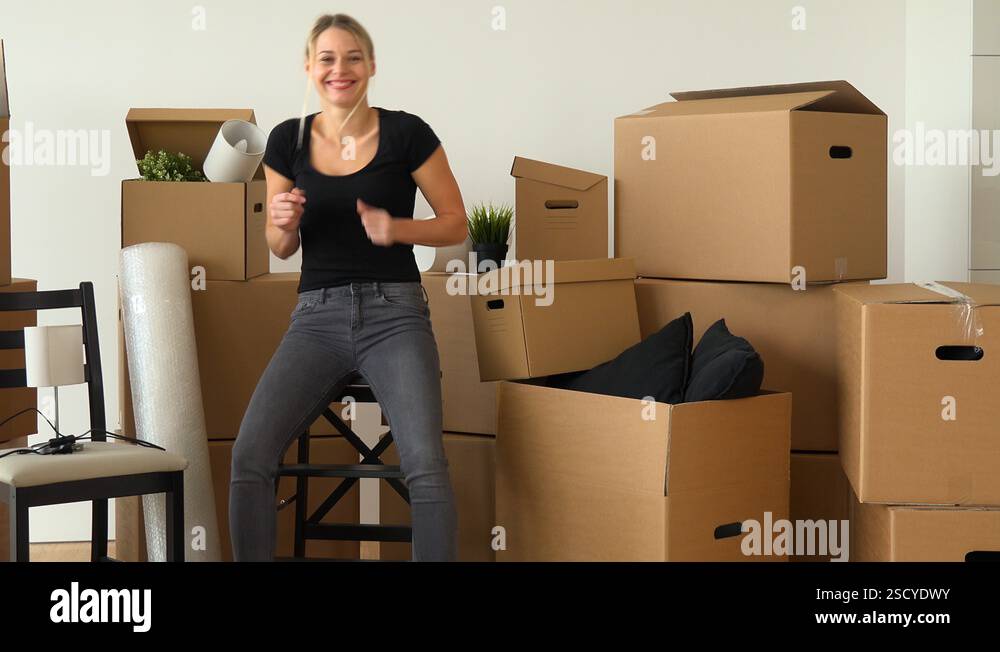 A happy moving woman sits on a chair in an empty apartment, dances and ...