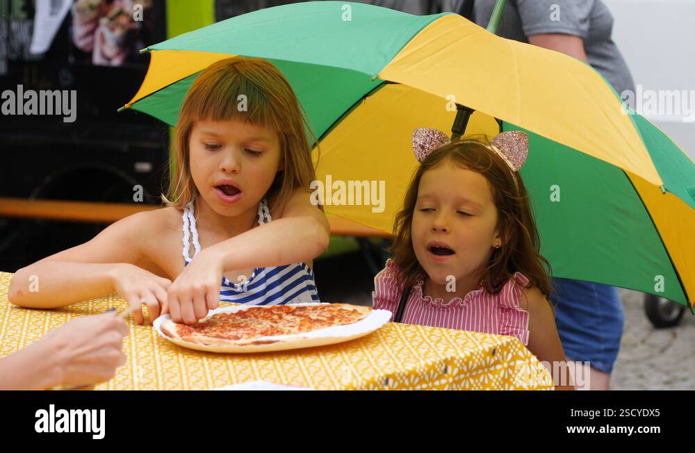 Two children girls eat street food pizza on festival rainy day under ...