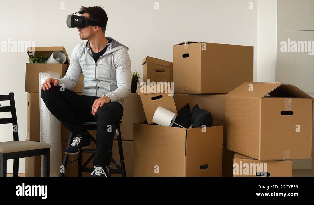 A moving man sits on a chair in an empty apartment and uses VR headset ...