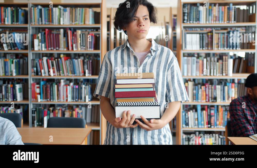 Serious teenage boy walking in library with bunch of books looking ...