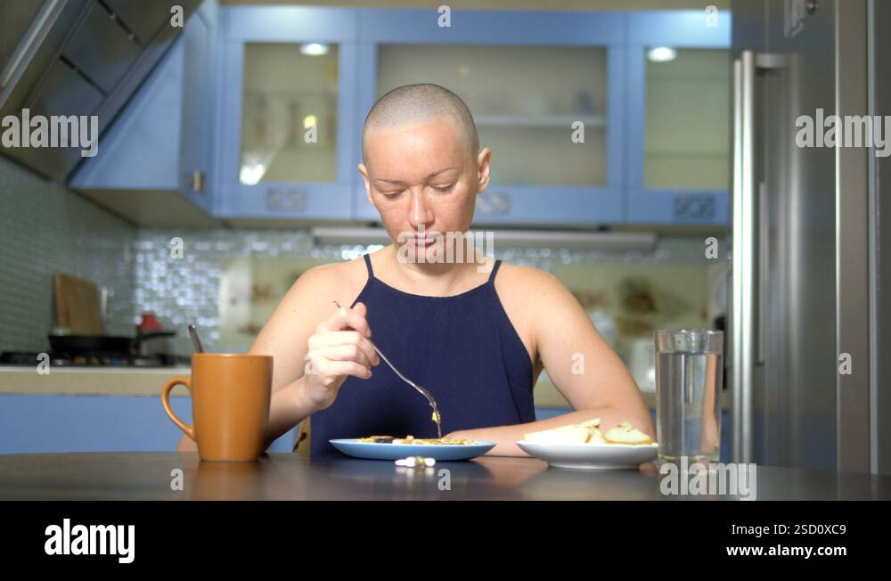 bald sad woman sits at a table in the kitchen with food and pills ...