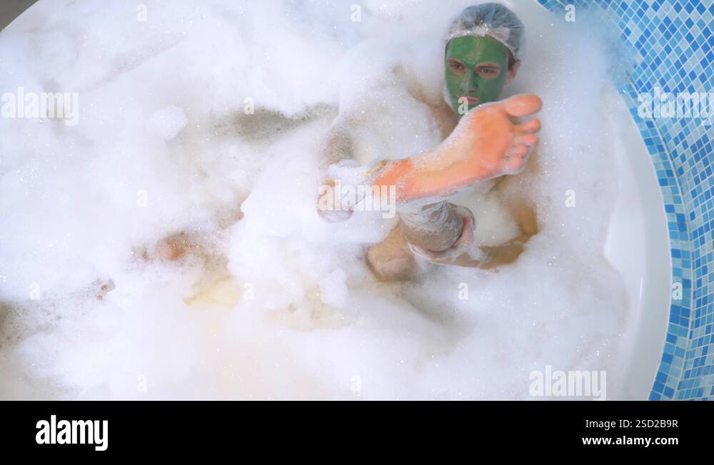 A man in a shower cap and a green face mask lies in a bath with copious ...