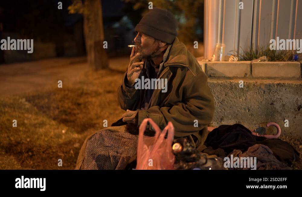 Lonely cold homeless man sitting on the street at night smoking ...