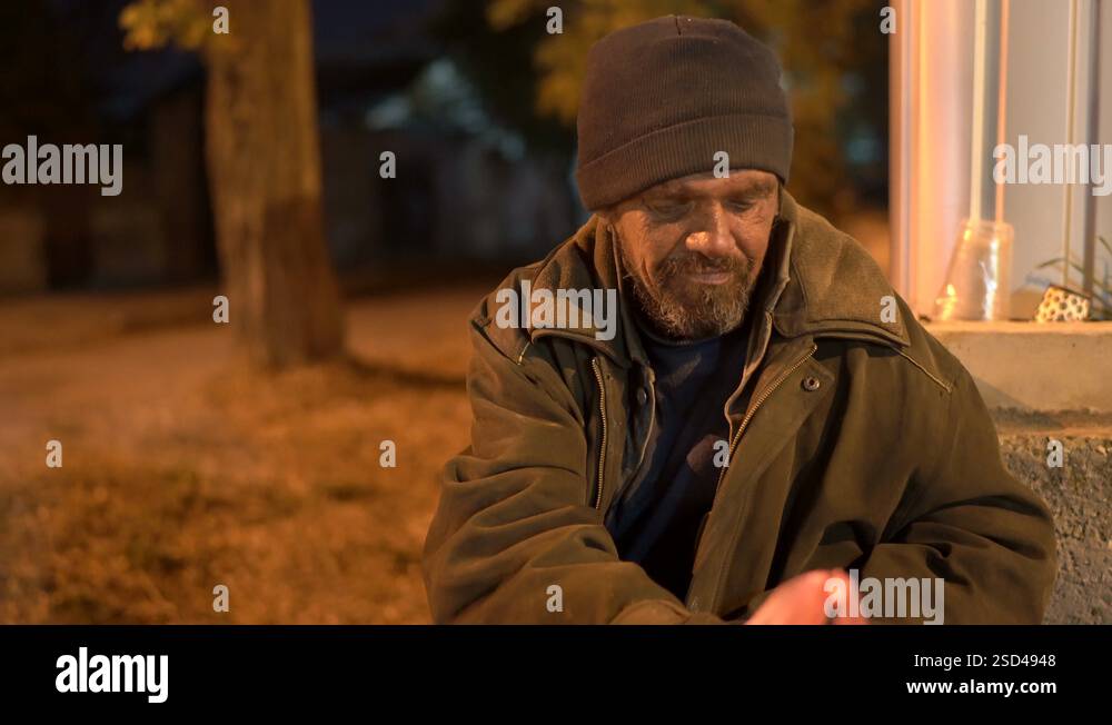Homeless sick man sitting on the street at night. Lonely cold beggar ...