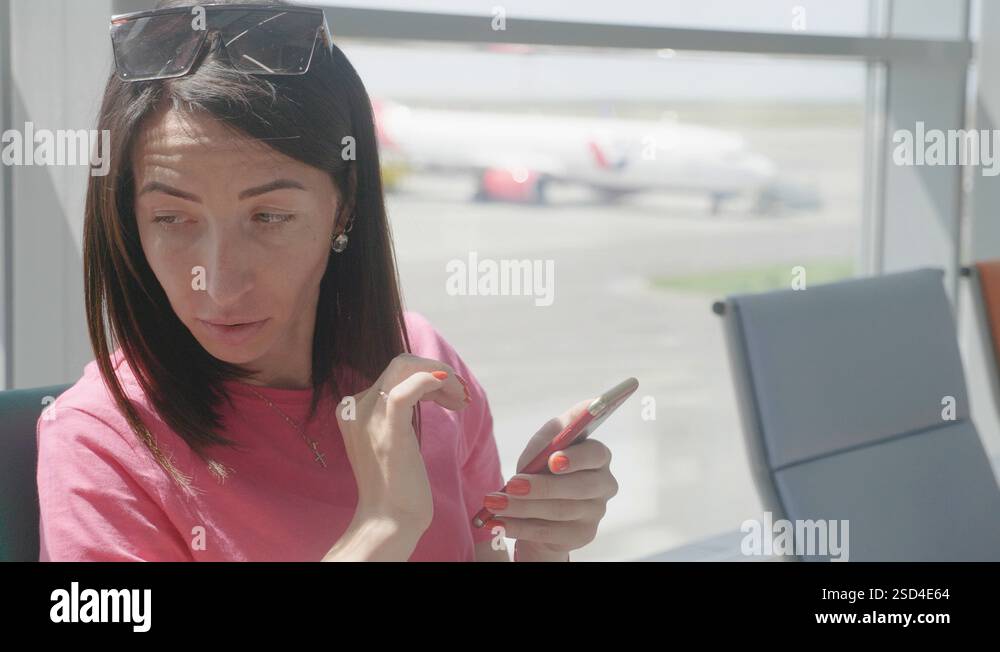 A young beautiful girl sits in the airport waiting room and uses a ...